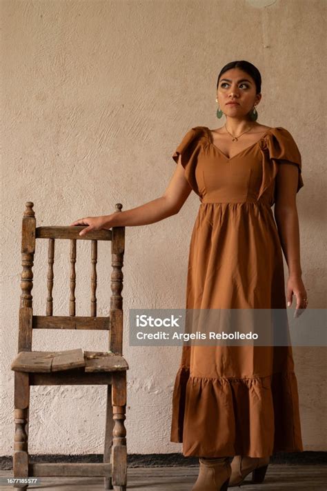 멕시코 여성이 섬세한 아치형 통로 아래 늙은 의자를 우아하게 장식하고 있다 빛과 그림자의 놀이에 흠뻑 젖어 있는 그녀의 전통 의상과 사색적인 시선 25 29세에 대한 스톡