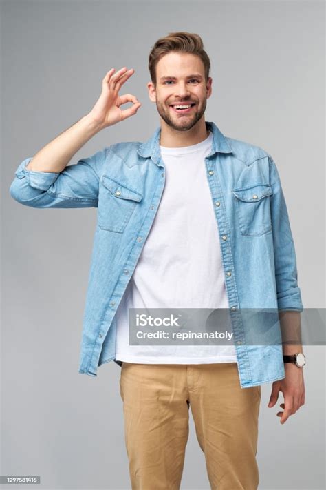 청바지 셔츠를 입은 젊은 잘생긴 백인 남자의 초상화가 밝은 배경 위에 서 있는 확인 사인 제스처를 보여줍니다 25 29세에 대한
