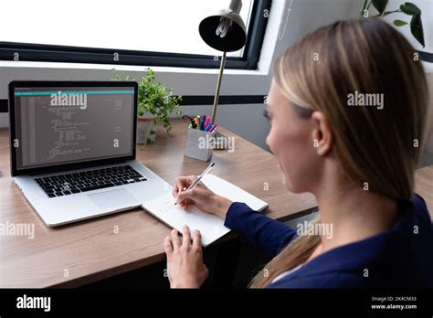 Caucasian Female Programmer Siting At Desk At Laptop With Coding On Screen Making Notes Coding