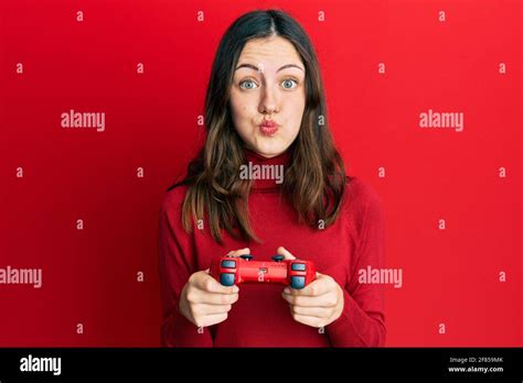 Young Brunette Woman Playing Video Game Holding Controller Puffing Cheeks With Funny Face Mouth
