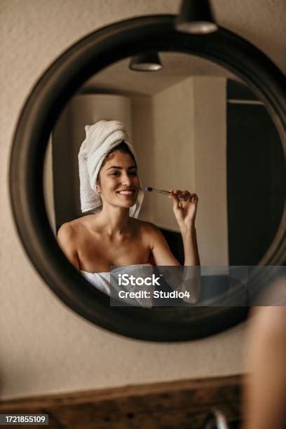 수건에 싸인 고요한 욕실의 라틴 여성은 양치질을 하면서 밝은 미소로 하루를 시작하고 눈은 거울에 고정되어 있습니다 25 29세에