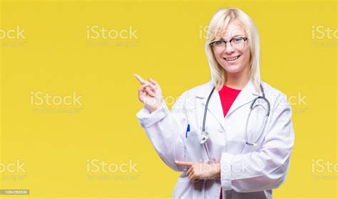 젊은 아름 다운 금발 의사 여자 얼굴에 큰 미소와 함께 고립 된 배경 위에 의료 유니폼을 입고 카메라를 보고 옆에 손 및 손가락으로 가리키는 가리키기에 대한 스톡 사진 및