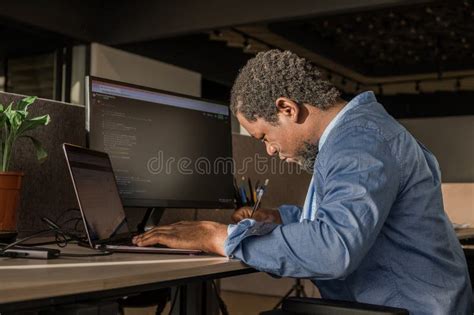 black man writing lines of code on desktop pc with two monitors and a