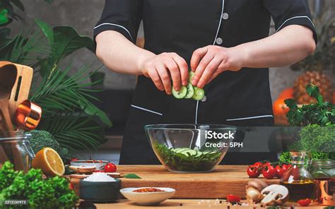 부엌에서 음식을 요리합니다 요리사는 얇게 썬 오이를 루꼴라와 함께 그릇에 뿌리고 샐러드를 준비합니다 주방용품 야채 허브 재료가 있는 나무 테이블에 건강한 비건 채식 요리법