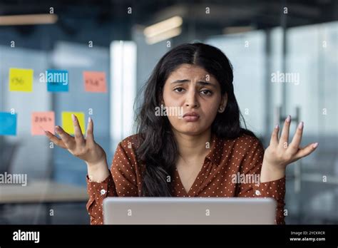 Confused Latina Businesswoman Shrugs Baffled At Desk Surrounded By