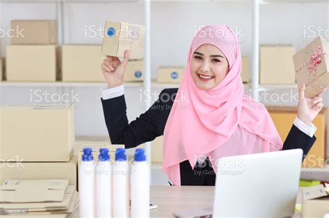 파란 양복을 입은 활동적인 아시아 무슬림 여성이 앉아서 컴퓨터로 일하고 행복한 웃는 얼굴로 온라인 소포 상자 배달을 보여주고