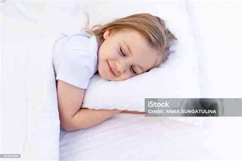 귀여운 아기 소녀가 그녀의 뺨 아래에 접힌 그녀의 손으로 베개를 껴안고 담요 아래 흰색 면 침대에 침대에서 자고 밤에 건강한 아기의 수면 6 7 살에 대한 스톡 사진 및