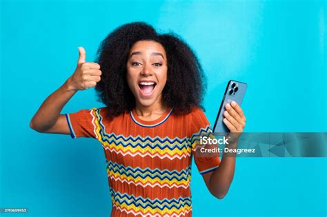 갈색 줄무늬 티셔츠를 입고 새로운 아이폰 15를 사용하여 파란색 배경에 고립 된 속도 엄지 손가락을 즐기는 젊은 재미 소녀 물결 모양의 헤어 스타일 사진 25 29세에 대한