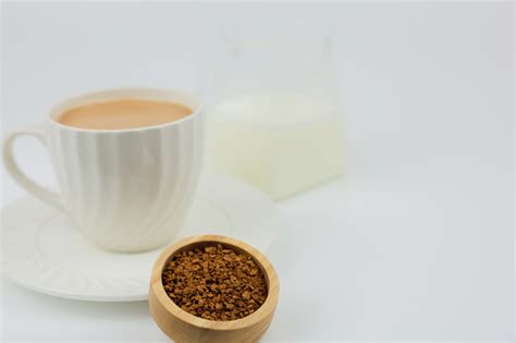 우유가 담긴 하얀 커피잔을 클로즈업한 사진이 접시 위에 놓여 있고 그 옆에는 텍스트를 넣을 수 있는 흰색 배경에 즉석 분쇄 커피와 우유 주전자가 담긴 나무 그릇이 있습니다