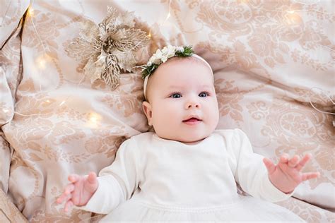 머리에 장식이 있는 흰색 드레스를 입은 어린 아기의 초상화를 클로즈업합니다 소녀는 빛의 화환에 밝은 소파에 누워 축제 크리스마스 트리 웃음 복사 공간 인사말 카드 아기에