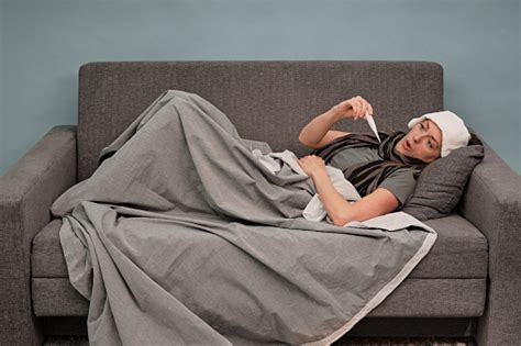 소녀는 집에서 아프다 소파에 앉아 담요에 싸여 목에 스카프를 두르고 있었다 전자 온도계로 온도를 측정합니다 코로나바이러스