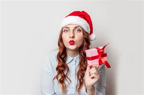 곱슬 머리와 범본과 빨간 모자와 산타 클로스 소녀는 빨간색 새틴 리본 활과 선물 상자를 보유하고 회색 벽의 배경에 카메라를보고 키스를 제공합니다 겨울에 대한 스톡 사진 및