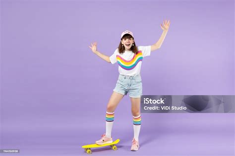 노란색 스케이트 보드와 함께 서 있는 생생한 옷을 입고 흥분 한 십 대 소녀가 스튜디오에서 보라색 파스텔 배경에 고립 된 손을 확산 시킵니다 사람들은 진심으로 감정 라이프