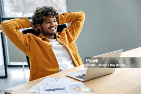 세련된 캐주얼 옷을 입은 긍정적 인 인도 남자 회사 직원은 업무용 책상에 앉아 노트북으로 일하면서 휴식을 취하고 머리 뒤로 손을 얹고 주말을 꿈꾸며 미소를 짓습니다 20