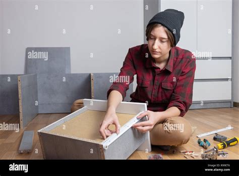 Woman Assembling A Chest Drawer For New Bedroom Furniture Female In Casual Shirt And Knit Hat