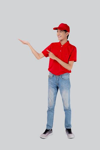 유니폼 빨간색과 모자를 쓴 젊은 아시아 남자는 흰색 배경 직원 또는 딜러 쇼 택배 및 배달 배달원 및 표현 물류 및화물에 고립 된 흥분으로 제시합니다 가리키기에 대한 스톡