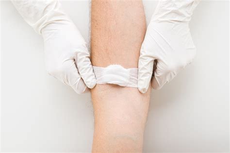 의사는 흰색 보호 고무 장갑을 끼고 밝은 회색 테이블 배경에 혈액 검사 후 젊은 성인 남자 팔 정맥에 목화솜으로 붕대를 감습니다 근접 촬영 환자 응급 처치 하향식 보기