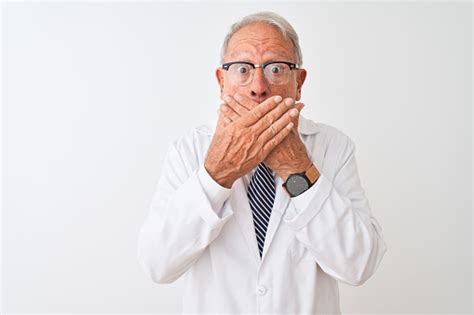 고립 된 흰색 배경 위에 서있는 코트를 입은 선임 회색 머리 과학자 남자는 실수로 손으로 입을 덮는 충격을 받았습니다 비밀 개념입니다 가리기에 대한 스톡 사진 및 기타