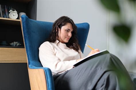 한 여자가 파란 의자에 앉아서 공책에 글을 쓰고 있다 그녀는 글쓰기에 집중하고 참여합니다 의자는 책장 앞에 있습니다 30 34세에 대한 스톡 사진 및 기타 이미지 Istock