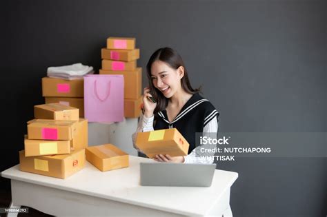 중소기업 사업주 긴 머리를 가진 젊은 아시아 여성 주문에 대해 고객과 전화 통화 사무실 테이블 위에 노트북이 있습니다 그 옆에는 보낼 준비가 된 소포 상자가 있습니다 25