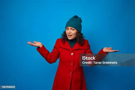깜짝 놀란 젊은 곱슬 머리 갈색 갈색 머리 여자 에 빨간 코트와 녹색 모자 포즈 에 푸른 배경에 대 한 포즈 그녀의 손 손바닥에 상상의 복사 공간을 들고 카메라를 보고