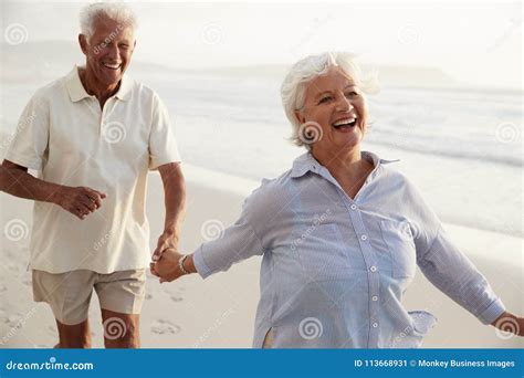 Senior Retired Couple Running Along Beach Hand In Hand Together Stock