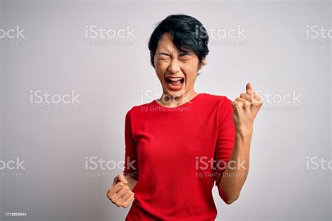 고립 된 흰색 배경 위에 서있는 캐주얼 한 빨간 티셔츠를 입은 젊은 아름다운 아시아 소녀는 팔을 들고 눈을 감고 성공을 축하하고 놀랐습니다 우승자 컨셉 T 셔츠에 대한 스톡