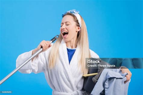 청소하는 동안 걸레 막대기에 노래 긍정적 인 행복한 여성은 바닥을 닦는 동안 주위를 바보로 만듭니다 그녀는 손에 양동이를 들고 푸른 스튜디오 배경에 포즈를 취합니다 가구에