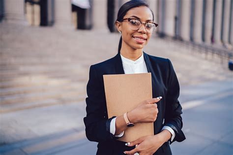 성공적인 미소 아프리카 계 미국인 여성 직원의 절반 길이 초상화 폴더를 들고 카메라에 미소 를 입고 세련된 양복을 입고 도시 설정에 서 긍정적 인 어두운 피부 사업가 거리에