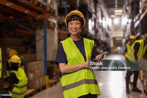 자신 있는 중성인 여성 창고 작업자가 안전 조끼와 안전모를 착용하고 디지털 태블릿을 사용하여 대형 물류 창고에서 재고 관리를 하면서 위를 올려다보고 있습니다 30 34세에