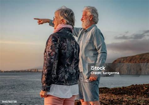바다의 바위 위에 석양이 비치는 곳에 서서 물 위의 수평선을 바라보는 평온한 노부부 여행과 자연을 즐기는 백인 은퇴자 부부를