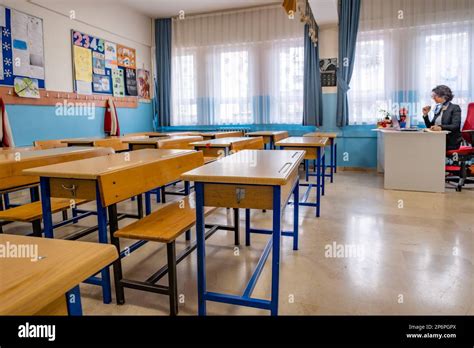 Empty Class Room With Wood Tables And Female Teacher Sitting At Her Desk Reading Notes In Her