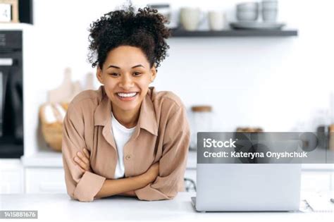 초상화 사랑스러운 긍정적 인 곱슬 머리 아프리카 계 미국인 젊은 여성 캐주얼 한 세련된 옷 여학생 또는 프리랜서 노트북 근처의 부엌에서 집에 서서 카메라를보고 행복하게 미소