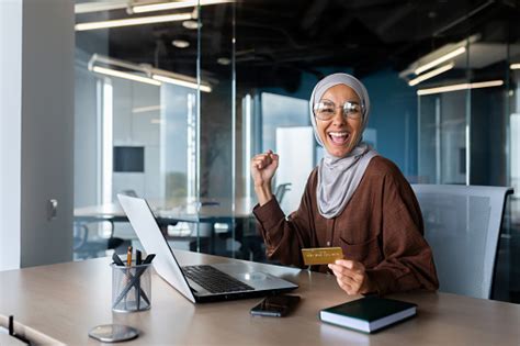 히잡을 쓴 행복한 무슬림 여성이 노트북을 들고 사무실에 앉아 신용 카드를 손에 들고 카메라를 향해 즐겁게 웃고 있습니다 30
