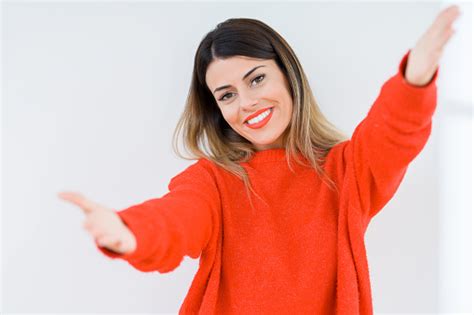 젊은 여성이 고립 된 배경 위에 캐주얼 빨간 스웨터를 입고 포옹을 위한 열린 팔로 웃 고 카메라를 보고 행복을 품은 쾌활 한 표현 갈색 머리에 대한 스톡 사진 및 기타