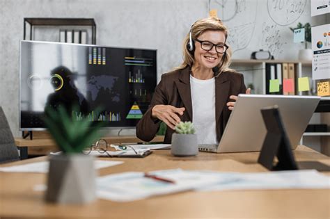 공식적인 옷과 헤드셋을 입은 아름다운 성숙한 백인 여성이 책상에 앉아 보고서를 작성합니다 40 44세에 대한 스톡 사진 및 기타 이미지 40 44세 가정의 방 경영자
