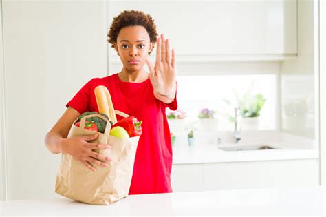진지하고 자신감있는 표정 방어 제스처로 정지 표지판을하는 오픈 손으로 신선한 식료품으로 가득 찬 종이 가방을 들고있는 젊은 아프리카 계 미국인 여성 가방에 대한 스톡 사진