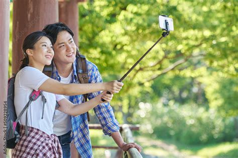 한국의 옛날 전통 가옥의 처마와 녹색 숲을 배경으로 여행하며 스마트폰으로 다양한 포즈를 셀카로 찍는 젊은 아시아 한국 남녀 모델 Stock Foto Adobe Stock