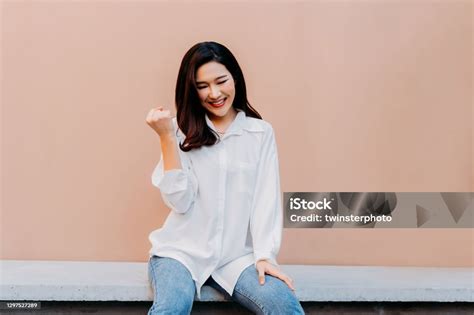 캐주얼 화이트 셔츠와 청바지 옷을 입고 배경에 사막 모래 색 벽콘크리트 벤치에 앉아 큰 상을 수상처럼 꽉 그녀의 주먹을 들고 젊은 아시아 소녀를 응원 20 29세에 대한