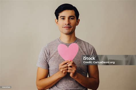 초상화 히스패닉 라티노 남자가 가슴 앞에 분홍색 하트 모양 종이를 들고 잘 생긴 젊은 성인 회색 티셔츠를 들고 카메라를 보며