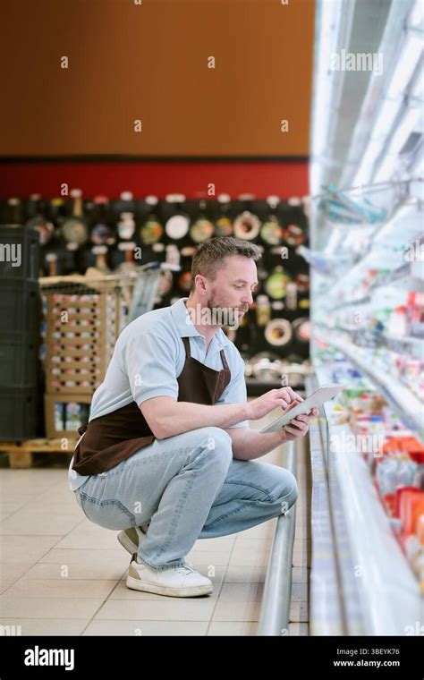 Employee Wearing Apron Crouching By Refrigerated Section In Grocery