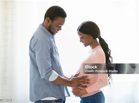 집에서 창 근처에서 아내의 임신 한 배를 만지는 잘 생긴 흑인 남자의 측면 보기 임신에 대한 스톡 사진 및 기타 이미지 임신 2명 가정 생활 Istock