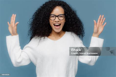 곱슬 머리와 혼합 레이스 여자를 기 뻐 손을 제기 긍정적 인 감정에서는 흰색 스웨터를 입고 입을 열어 유지 파란색 배경에 대 한 의미 얼굴 표정 개념 20 24세에 대한