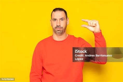 수염을 기른 잘생긴 히스패닉 남자는 손가락과 카메라로 작은 크기의 기호를 만드는 손으로 몸짓을 하는 빨간 스웨터입니다 측정 개념 노란색 배경에 격리 개념에 대한 스톡 사진