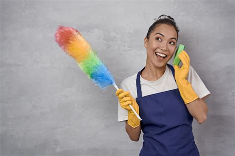 재미와 함께 청소 화려한 마이크로 화이버 더스터와 부엌 스폰지를 들고 유니폼을 입은 장난 꾸러기 젊은 여성은 회색 벽에 서서 휴대 전화로 말하는 척합니다 25 29세에 대한