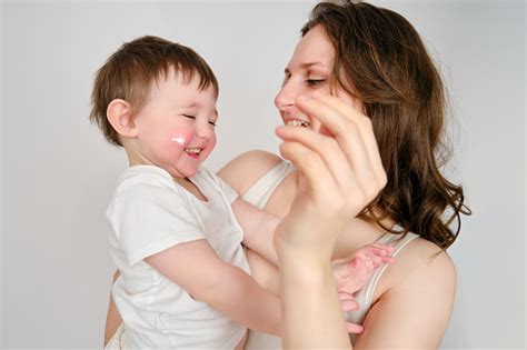 여자 어머니는 스튜디오 흰색 배경에 베이비 크림으로 뺨을 칠한다 엄마는 습진에 대한 보습제로 아이의 얼굴을 닦습니다 2세 무렵의 아이 보습제에 대한 스톡 사진 및 기타