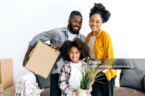 행복하고 즐거운 아프리카계 미국인 아빠 엄마와 유치원 귀여운 딸 새 아파트로 이사 집 물건이 담긴 상자를 들고 화분을 들고 있는