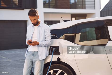 집 근처에서 차량 충전 스마트폰을 들고 젊은 세련된 남자가 낮에 전기 자동차와 함께 전기 자동차 대체 연료 자동차에 대한 스톡 사진 및 기타 이미지 전기 자동차 대체