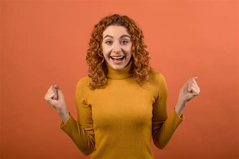 오렌지 색 스튜디오 배경에 고립 된 꽉 주먹으로 우승자 제스처를 보여주는 오렌지 점퍼에 젊고 매력적인 빨간 머리 백인 소녀 Holy Saturday에 대한 스톡 사진 및