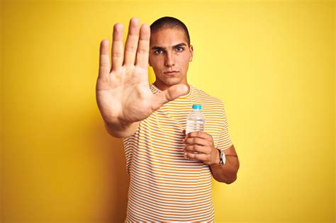 노란색 고립 된 배경 위에 플라스틱 물병을 들고 있는 젊은 잘생긴 남자는 진지하고 자신감있는 표정 방어 제스처로 정지 표지판을하는 오픈 핸드로 건강한 생활방식에 대한 스톡